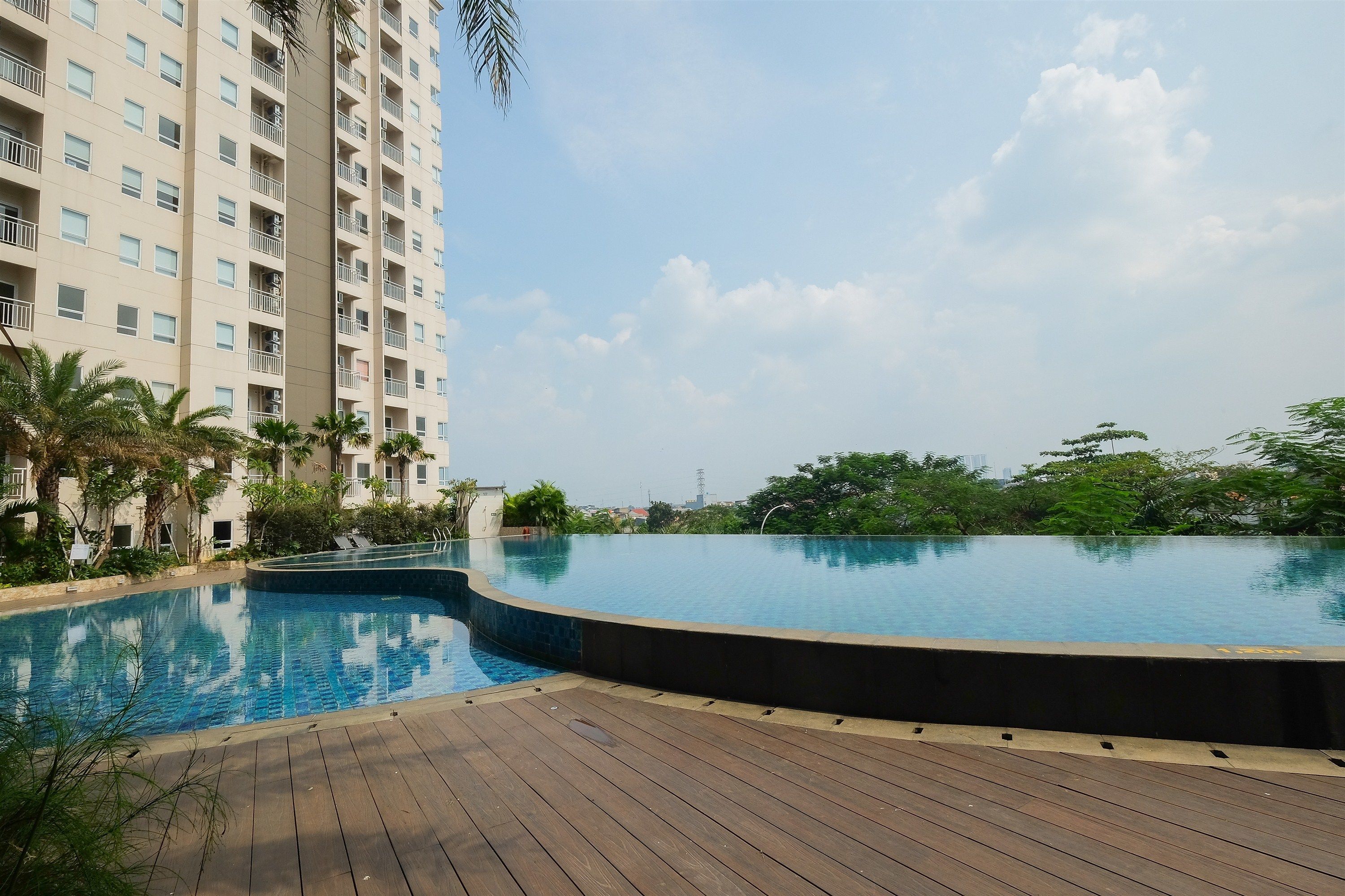Uma piscina com deck de madeira, rodeada por vegetação e um edifício alto ao fundo.