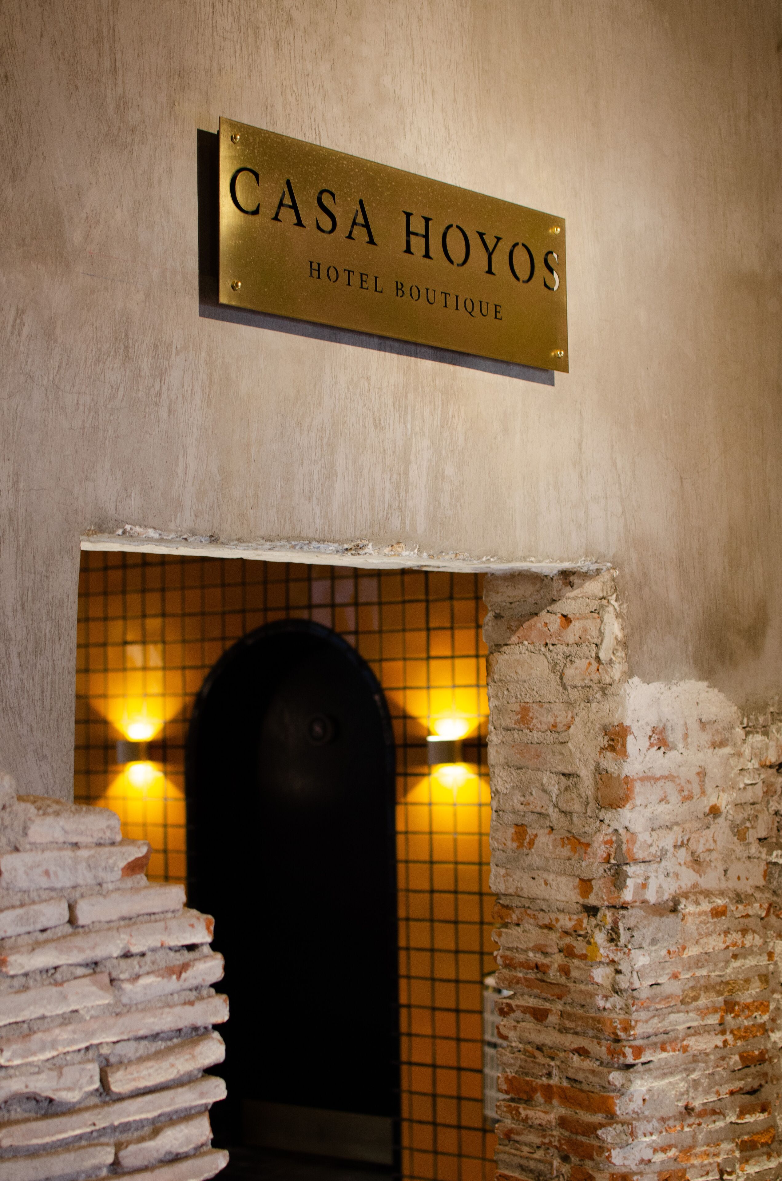 Placa com o nome 'CASA HOYOS HOTEL BOUTIQUE' acima de uma entrada, com parede de azulejos e estrutura de tijolos à direita.