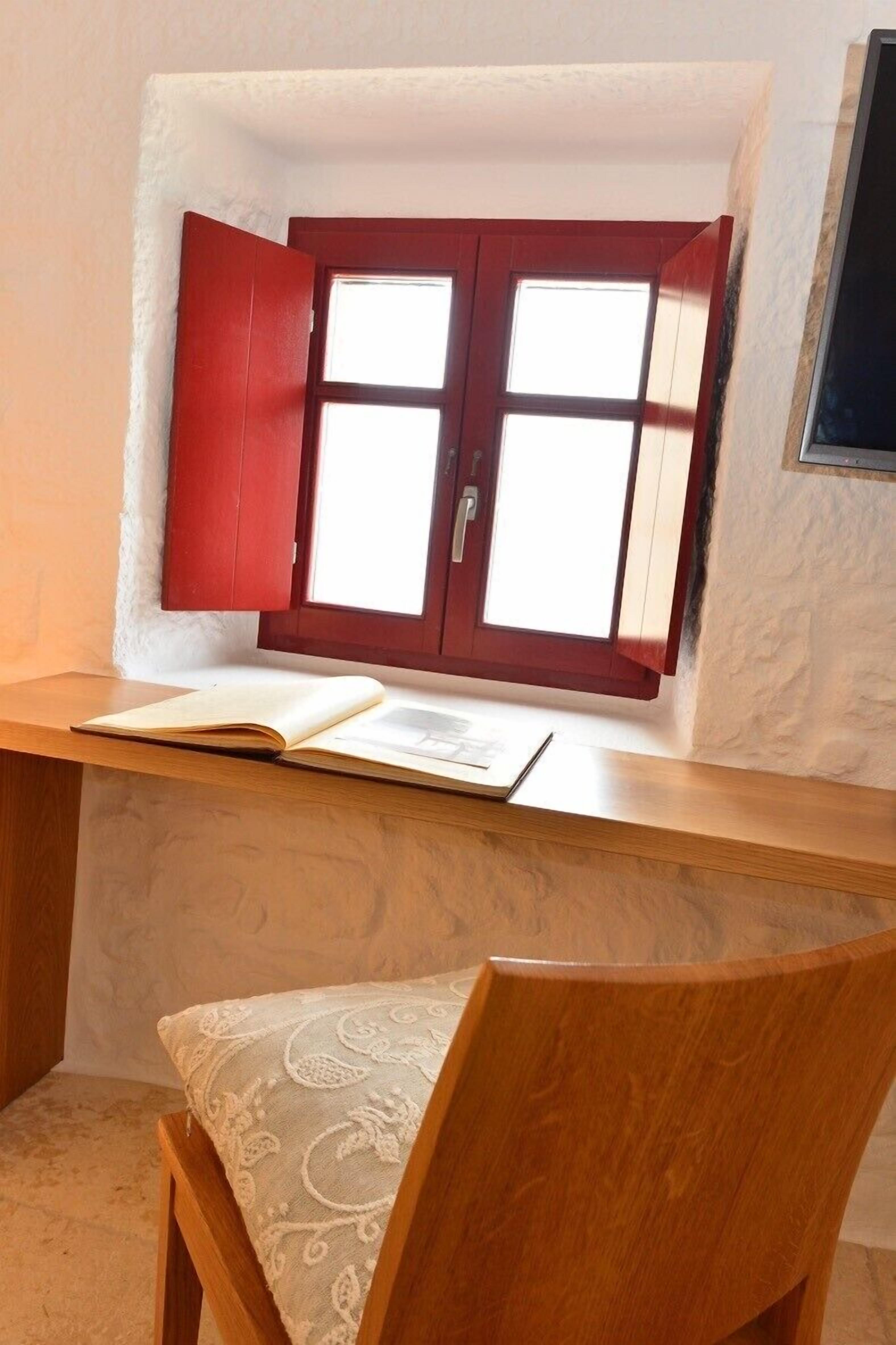 Mesa no canto com um armário vermelho, cadeira com almofada estampada e janela com cortinas brancas.