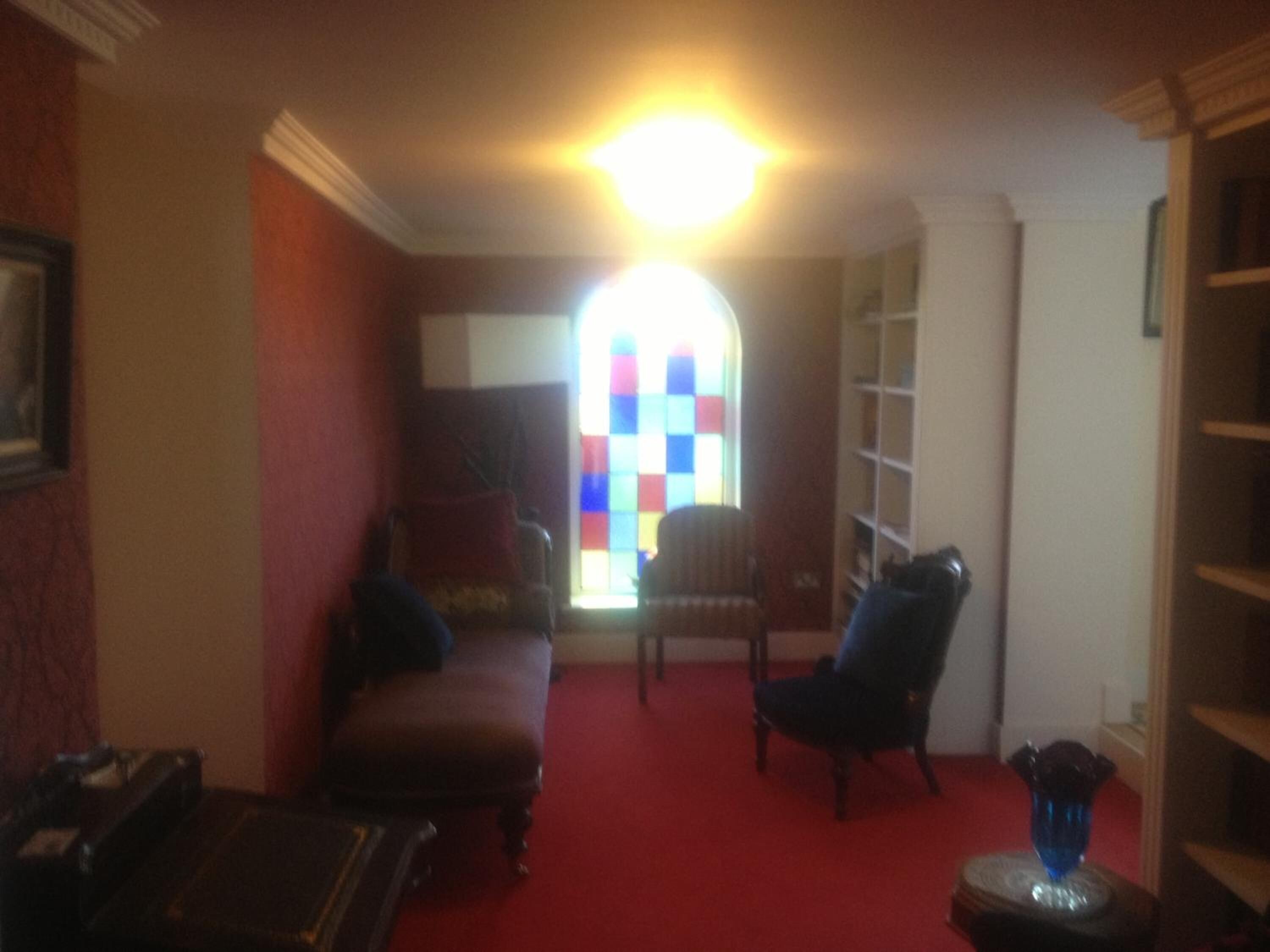 Um quarto acolhedor com carpete vermelho, uma janela grande com um desenho vibrante em vitral, uma estante e uma cadeira.