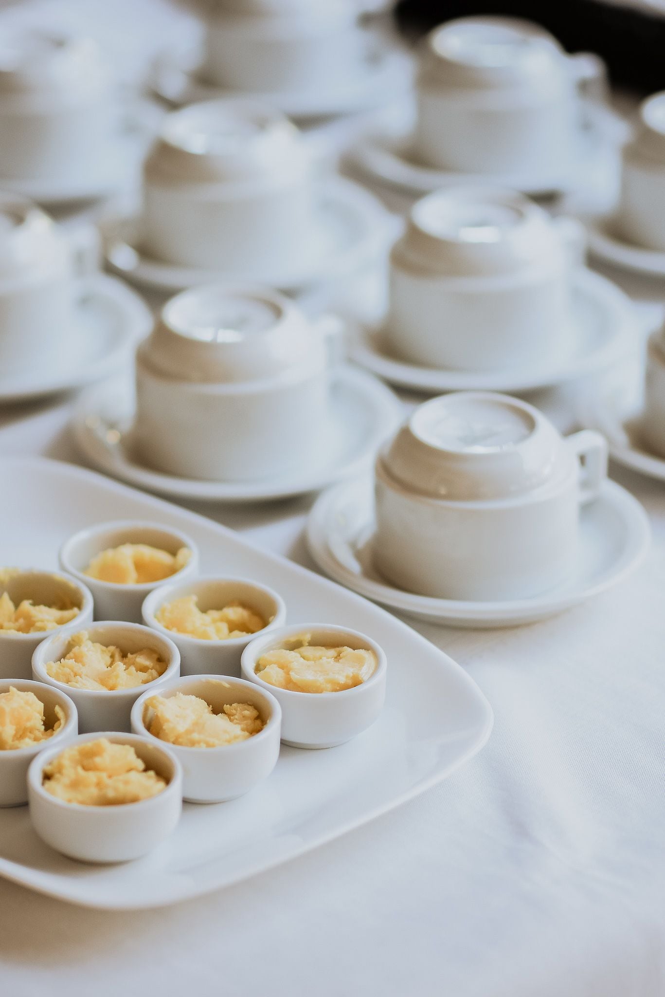 Uma bandeja com pequenas xícaras brancas e uma substância cremosa, possivelmente alimento, além de várias xícaras brancas maiores com tampas, provavelmente para bebidas.