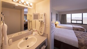 Quarto de hotel com banheiro equipado com pia, espelho e secador de cabelo, e duas camas, com janela que oferece uma vista.