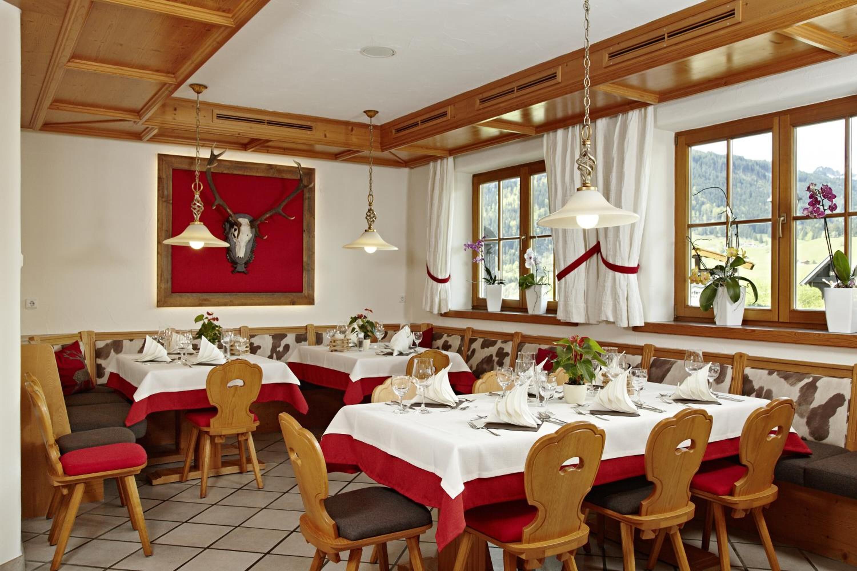Um restaurante com mesas e cadeiras de madeira, toalhas de mesa vermelhas e brancas, e uma vista panorâmica para as montanhas através de grandes janelas.