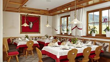 Um restaurante com mesas e cadeiras de madeira, toalhas de mesa vermelhas e brancas, e uma vista panorâmica para as montanhas através de grandes janelas.