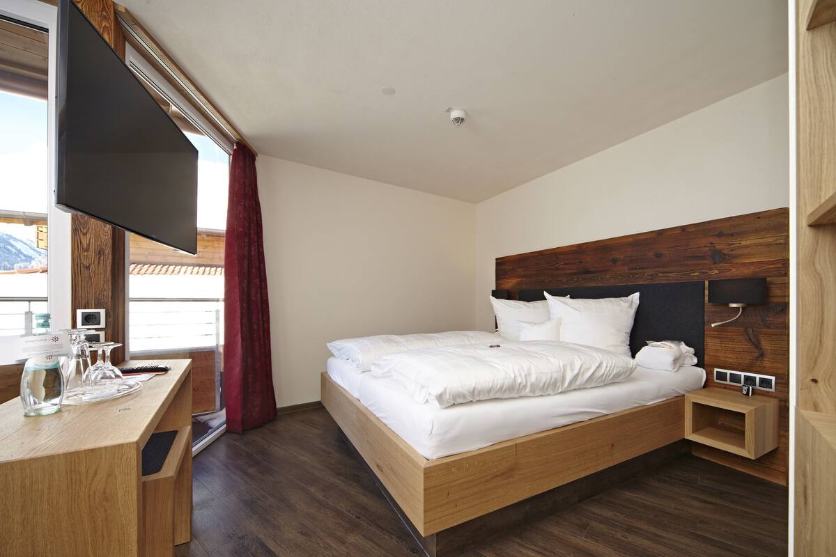 Quarto de hotel moderno com cama de madeira, escrivaninha com televisão de tela plana e vista para montanhas pela janela.