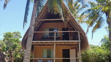 Uma casa com telhado de palha e varanda, cercada por palmeiras e vegetação.