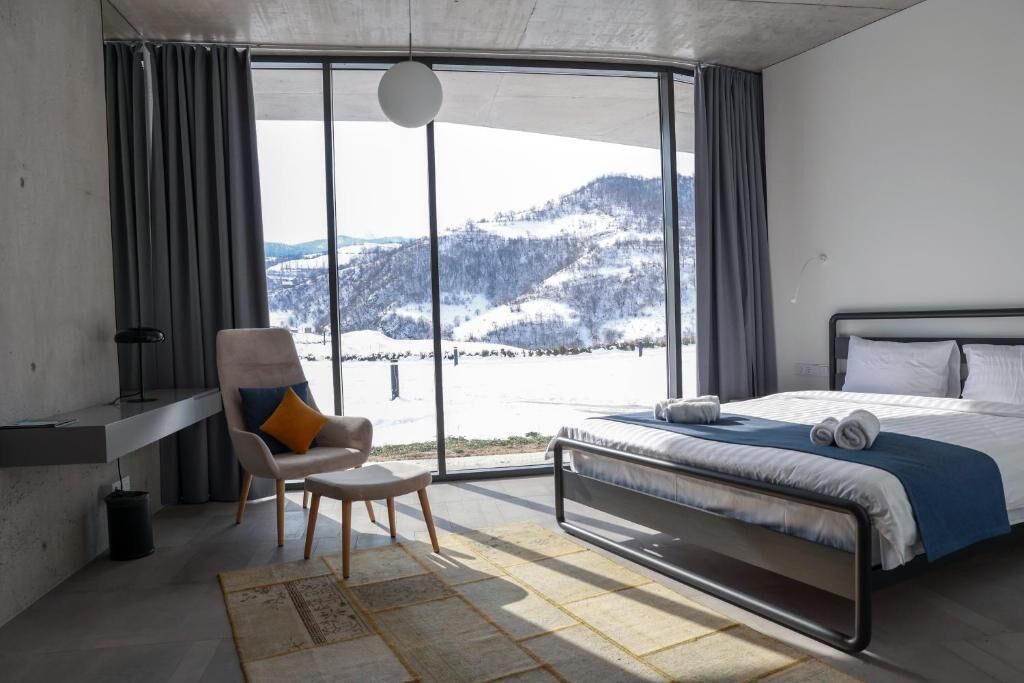 Quarto de hotel moderno com uma cama grande, uma cadeira, uma mesa e vista para colinas cobertas de neve.
