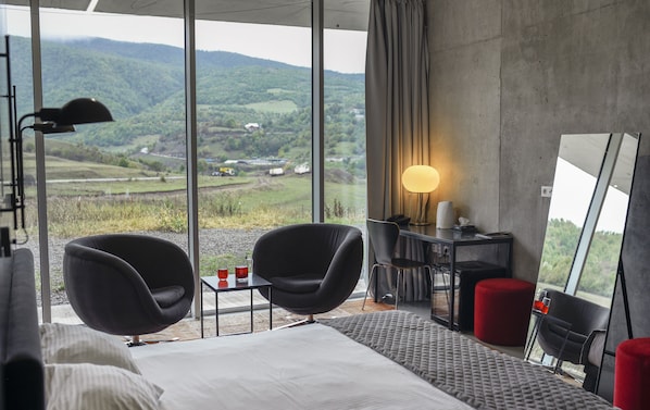 Quarto de hotel moderno com uma cama grande, duas cadeiras pretas, uma escrivaninha com abajur e vista para colinas verdes.