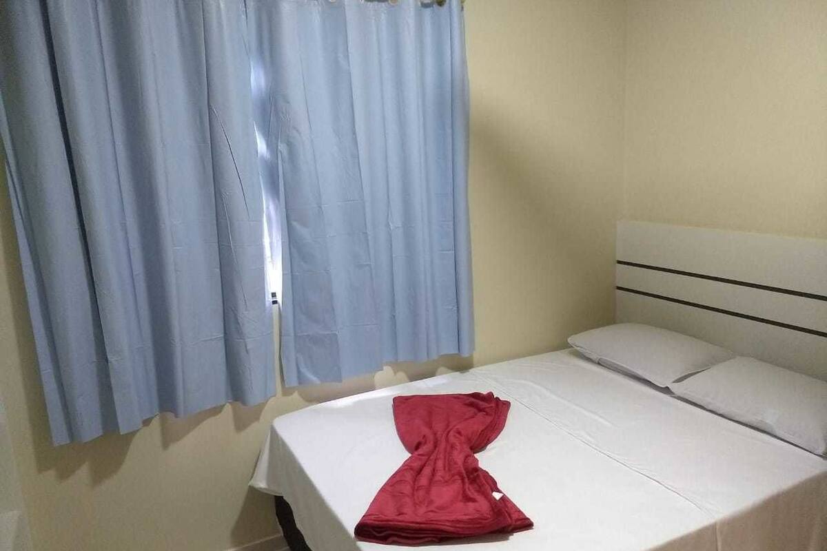 Uma cama de solteiro com colchão e cabeceira brancos, uma peça de roupa vermelha sobre a cama, e uma cortina azul parcialmente aberta.