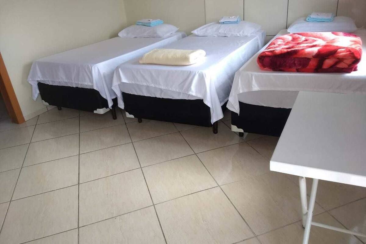 Quarto com três camas de solteiro, cada uma com colcha e travesseiros brancos. Há uma mesa branca em primeiro plano.