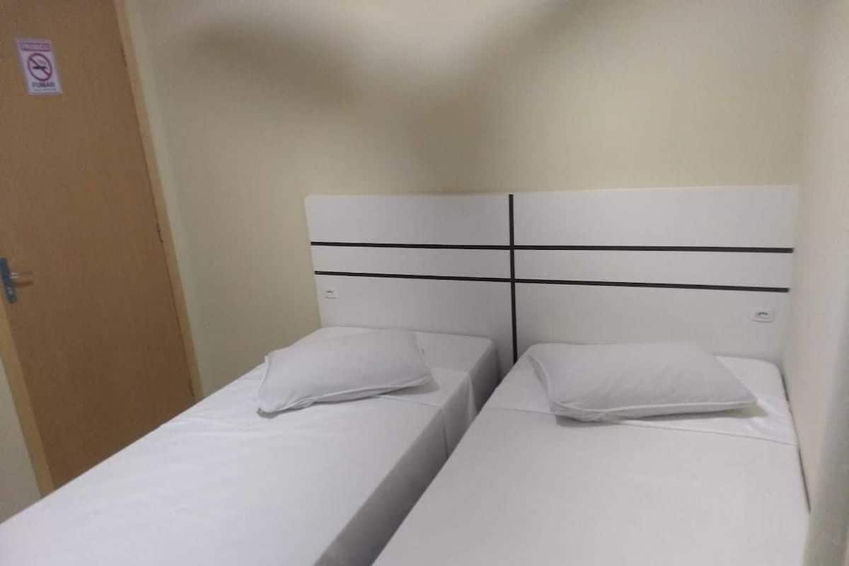 Quarto pequeno e simples com duas camas, cada uma com colchão e travesseiro brancos.
