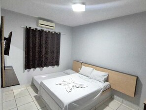 Uma cama de solteiro com roupa de cama branca num quarto com paredes cinzentas claras, uma cortina escura e um aparelho de ar condicionado sobre a cama.