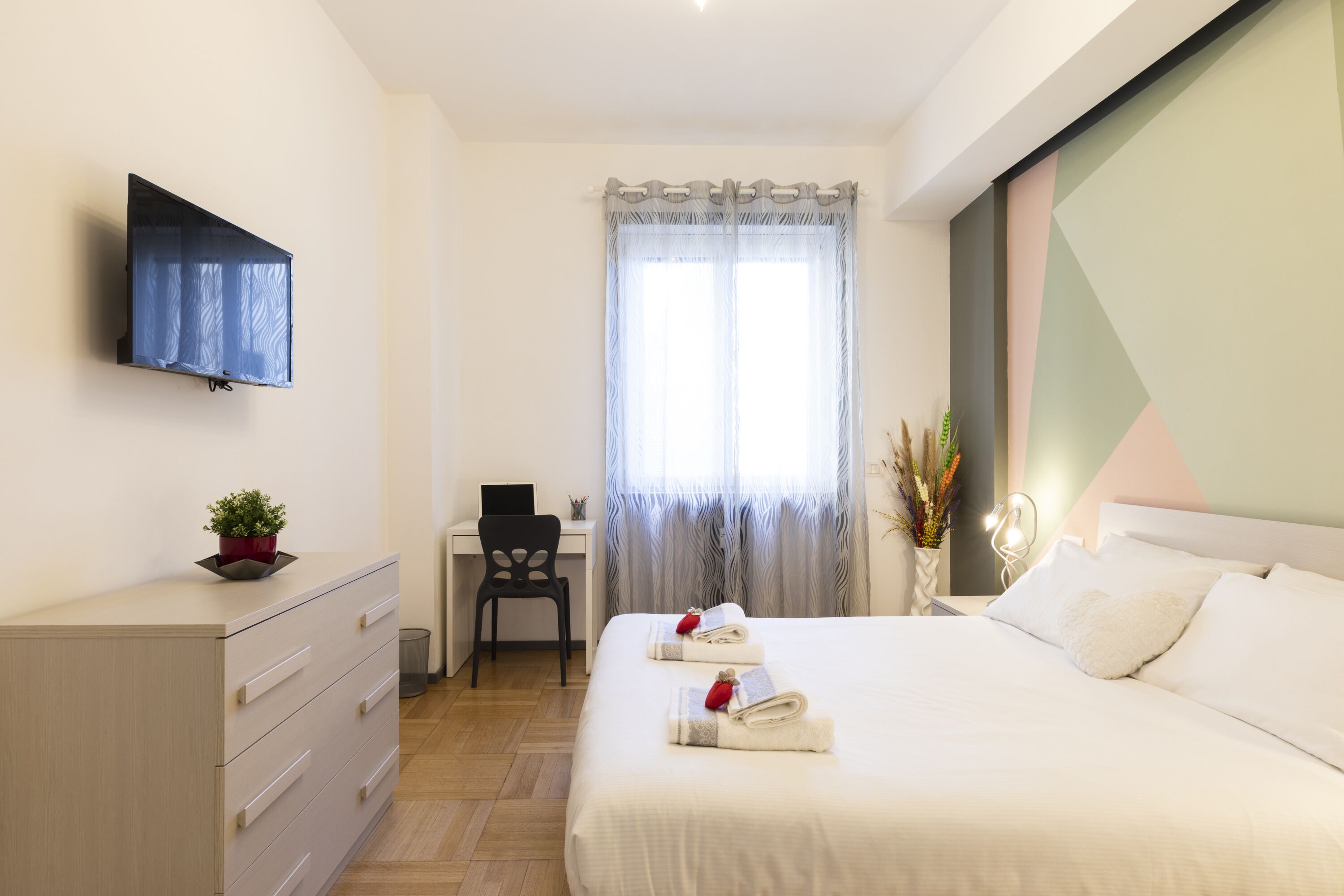 Quarto de hotel bem arrumado com cama, escrivaninha, cadeira, TV fixada na parede e janela com cortinas.
