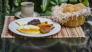 Prato de café da manhã com ovos, salsicha e torradas, uma xícara de café e uma cesta de pão em uma mesa de vidro com jogo americano listrado.