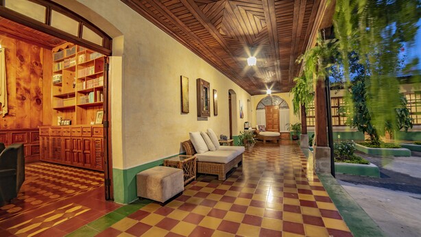 Um hall amplo com piso xadrez, estantes de madeira, área de estar com sofá e banquinho, e vista para o exterior.