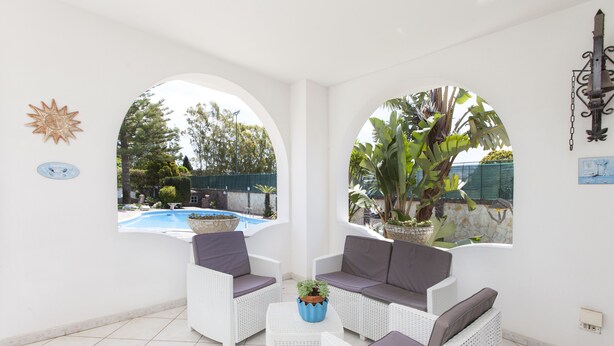 Pátio iluminado pelo sol com móveis de vime branco, uma pequena mesa com uma planta e vista para uma piscina e vegetação.