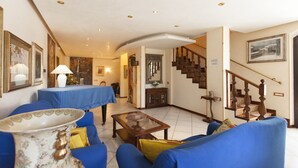 Sala de estar com sofás azuis, uma mesa de centro e uma mesa de jantar com toalha de mesa azul. Há uma escada com guarda-corpos de madeira e um lustre.