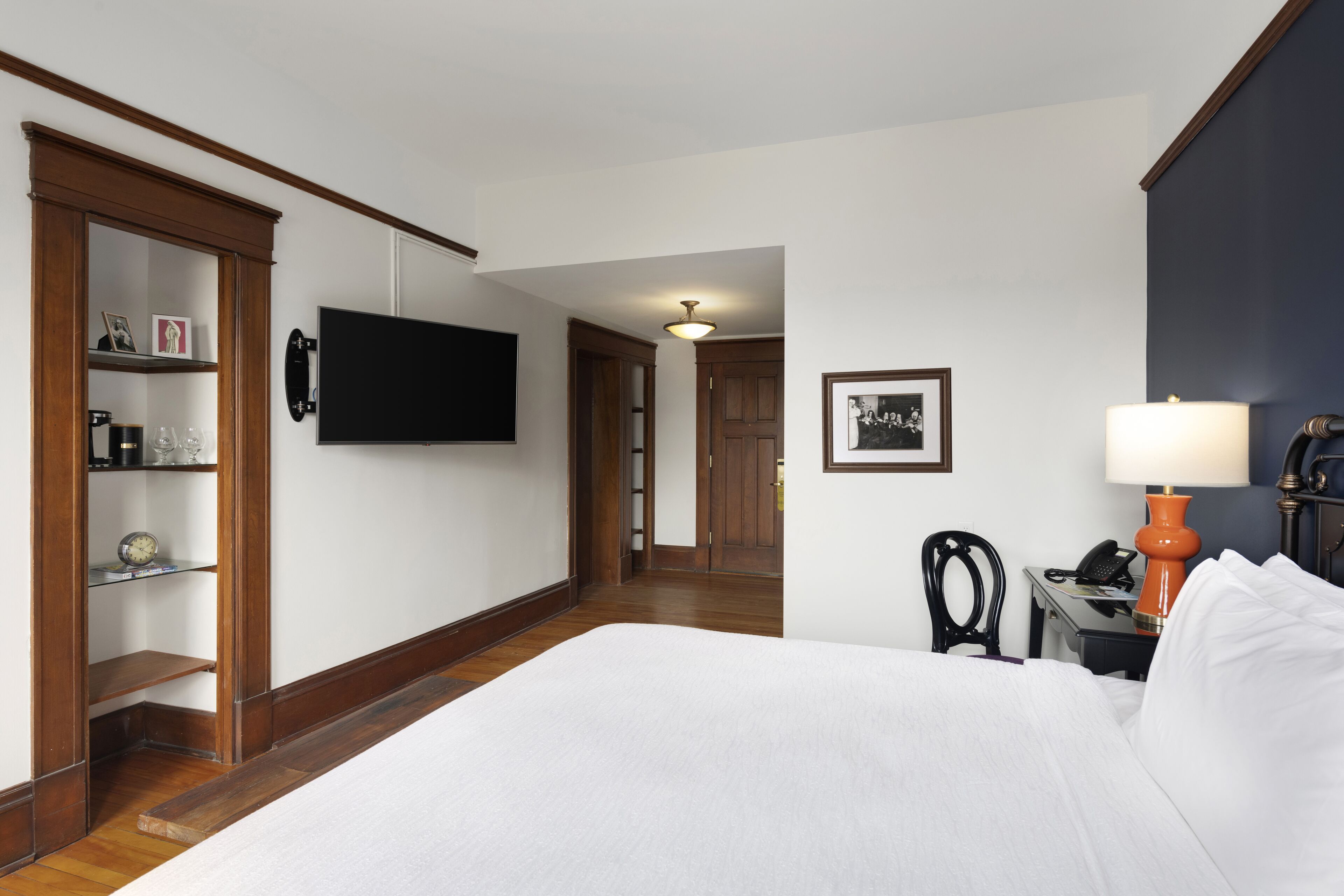 Quarto de hotel com uma cama grande, televisão na parede, escrivaninha com abajur e uma cadeira.