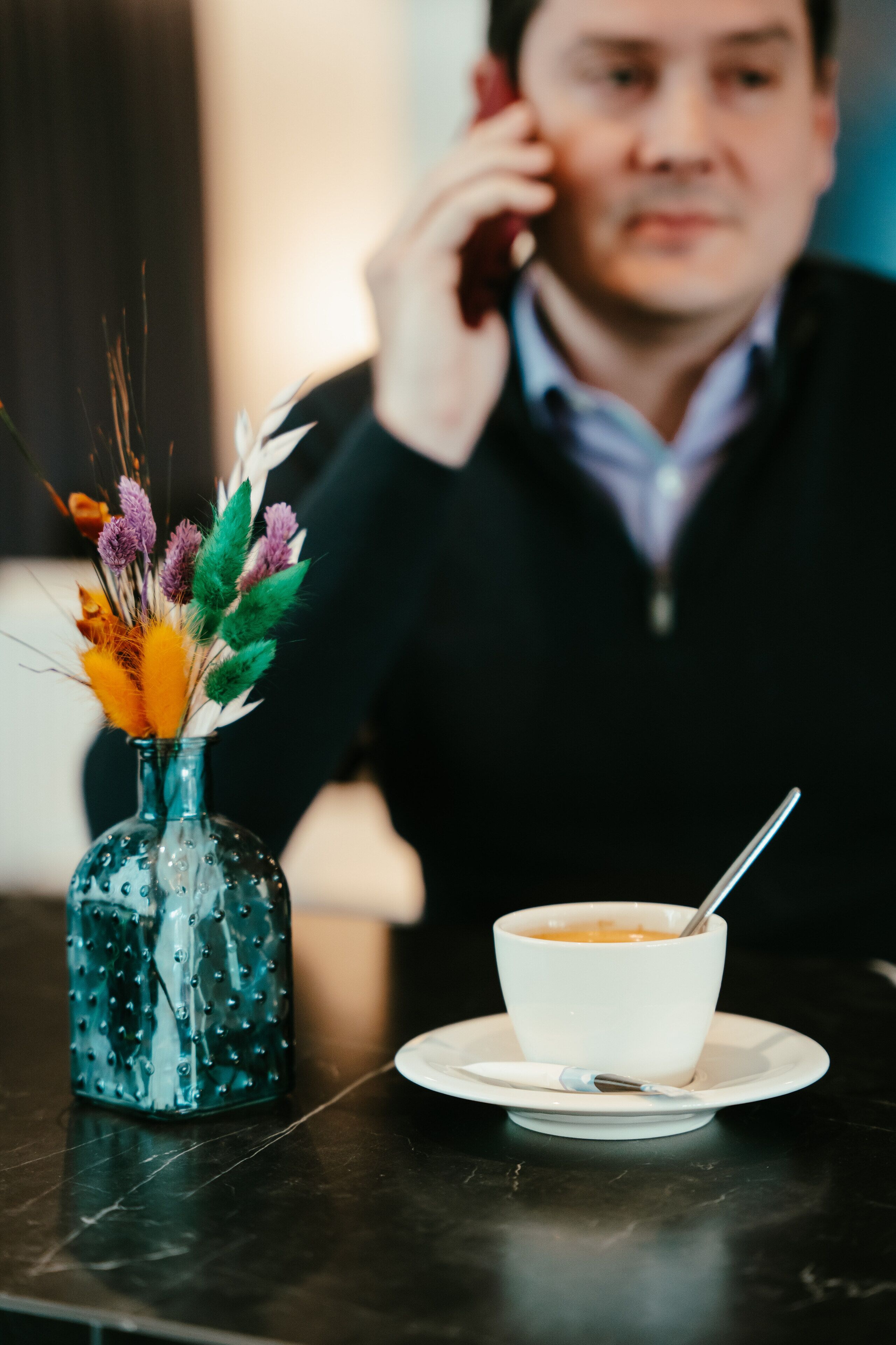 Uma pessoa sentada à mesa com uma xícara de café e um vaso de flores secas.