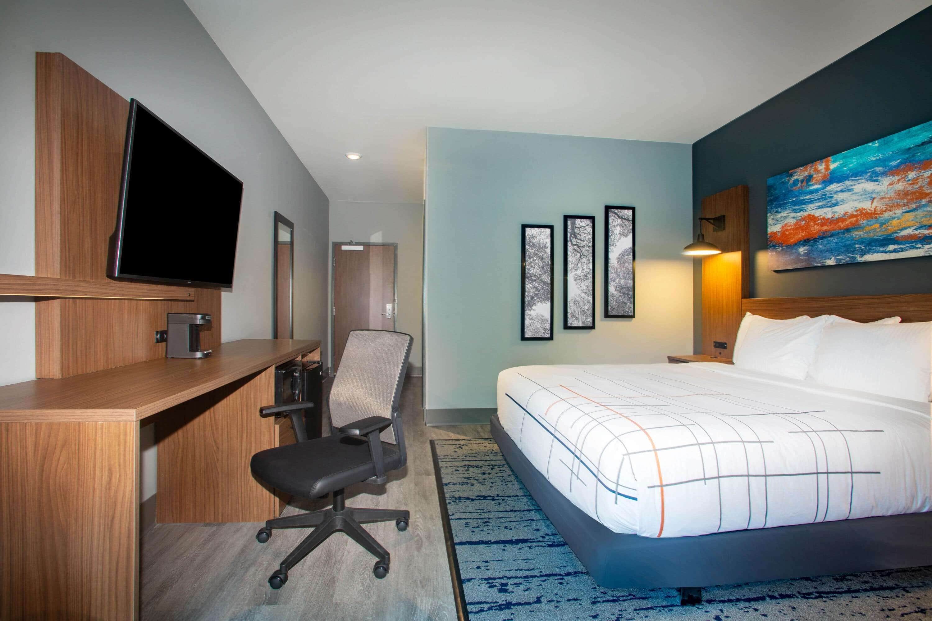 Quarto de hotel com uma cama, uma escrivaninha com cadeira, televisão de tela plana e quadros na parede.