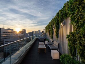 Terraço no telhado com guarda-corpo de vidro, área de estar ao ar livre e vista para o horizonte da cidade ao pôr do sol.