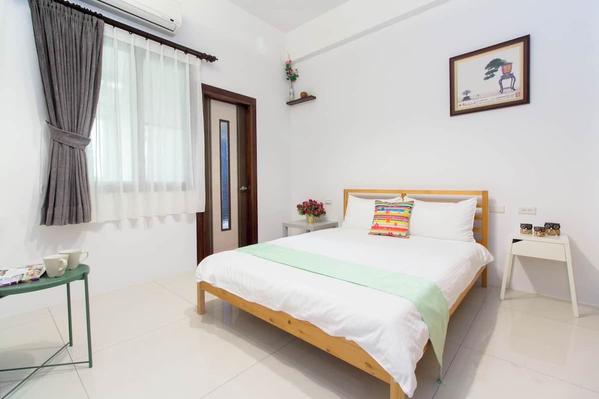 Um quarto arrumado com uma cama, mesas de cabeceira, uma pequena mesa com xícaras e uma janela com cortinas.