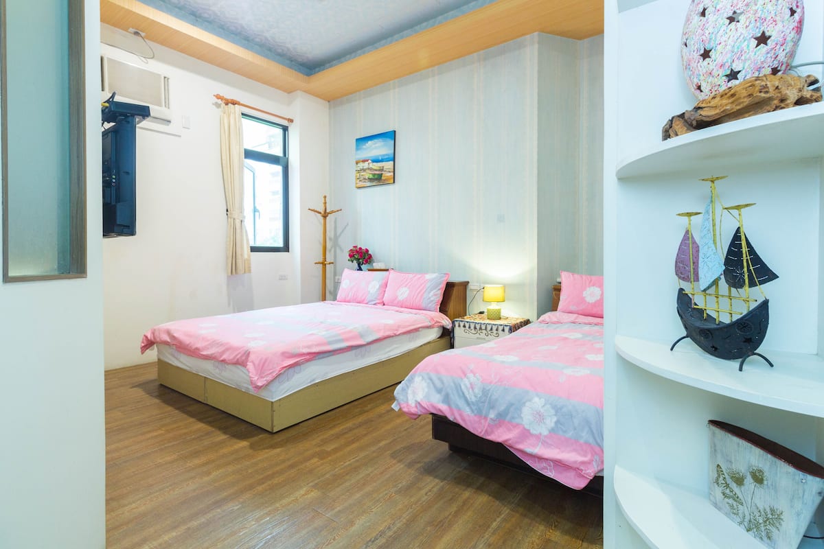 Quarto com duas camas, piso de madeira, uma janela, aquecedor fixado na parede e uma prateleira com objetos decorativos.