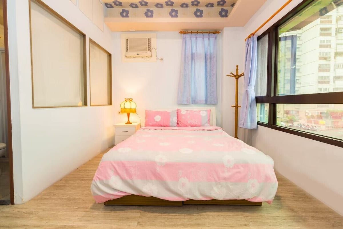 Quarto com cama, mesa de cabeceira, abajur, janela com cortinas e ar condicionado.