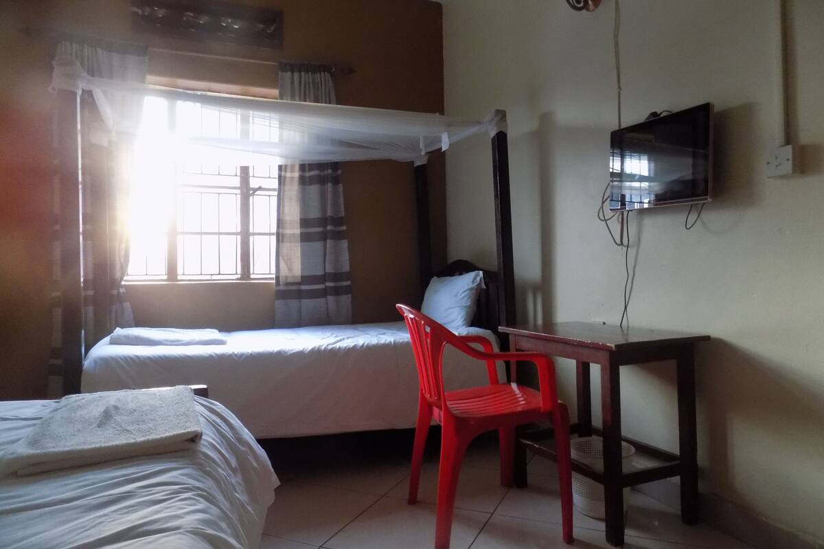Quarto com cama, escrivaninha, cadeira, televisão e janela com cortinas.