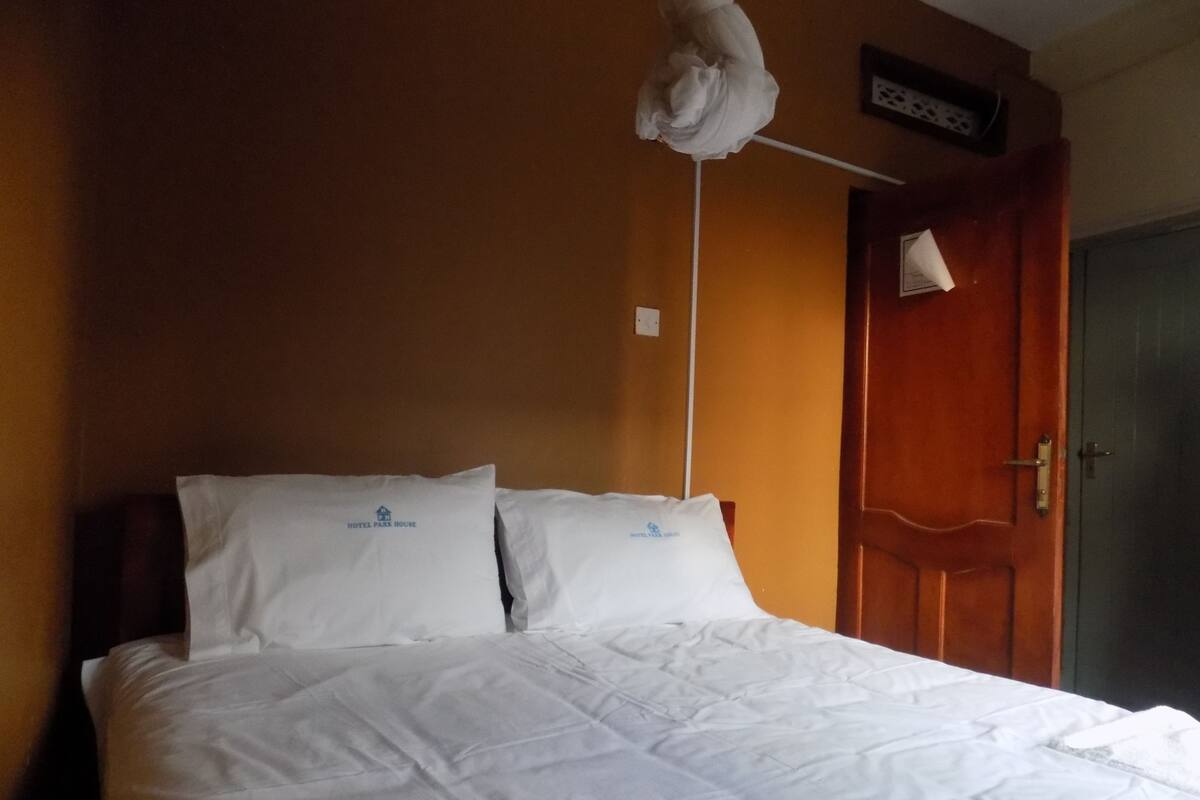 Quarto de hotel com cama, almofadas, porta de madeira e luminária fixada na parede.