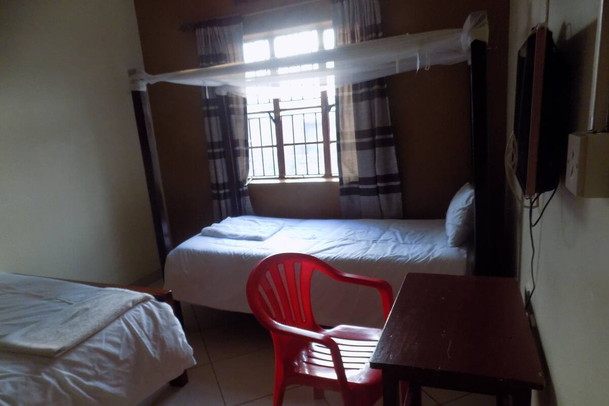 Quarto com duas camas, uma cadeira vermelha, uma mesa e uma janela com cortinas.