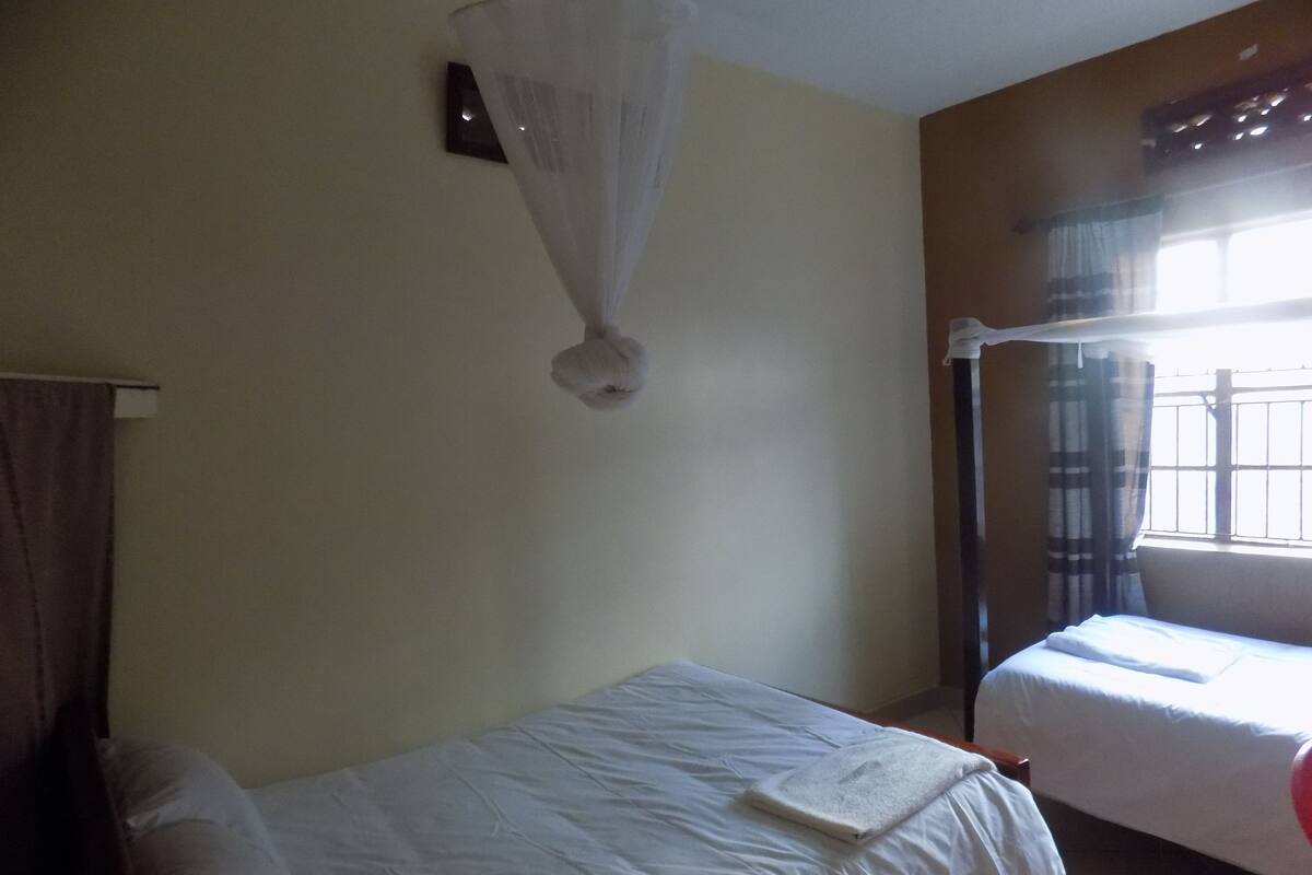 Quarto com duas camas, mosquiteiro, janela com cortinas e luminária na parede.