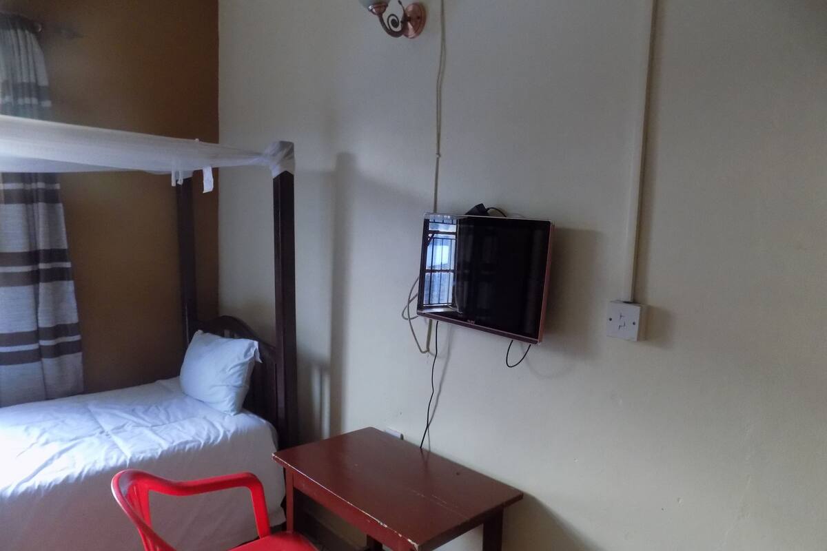 Uma cama de solteiro com colchão branco e uma cadeira de plástico vermelha. Uma pequena mesa de madeira está posicionada em frente à cama. Uma televisão está fixada na parede acima da mesa.