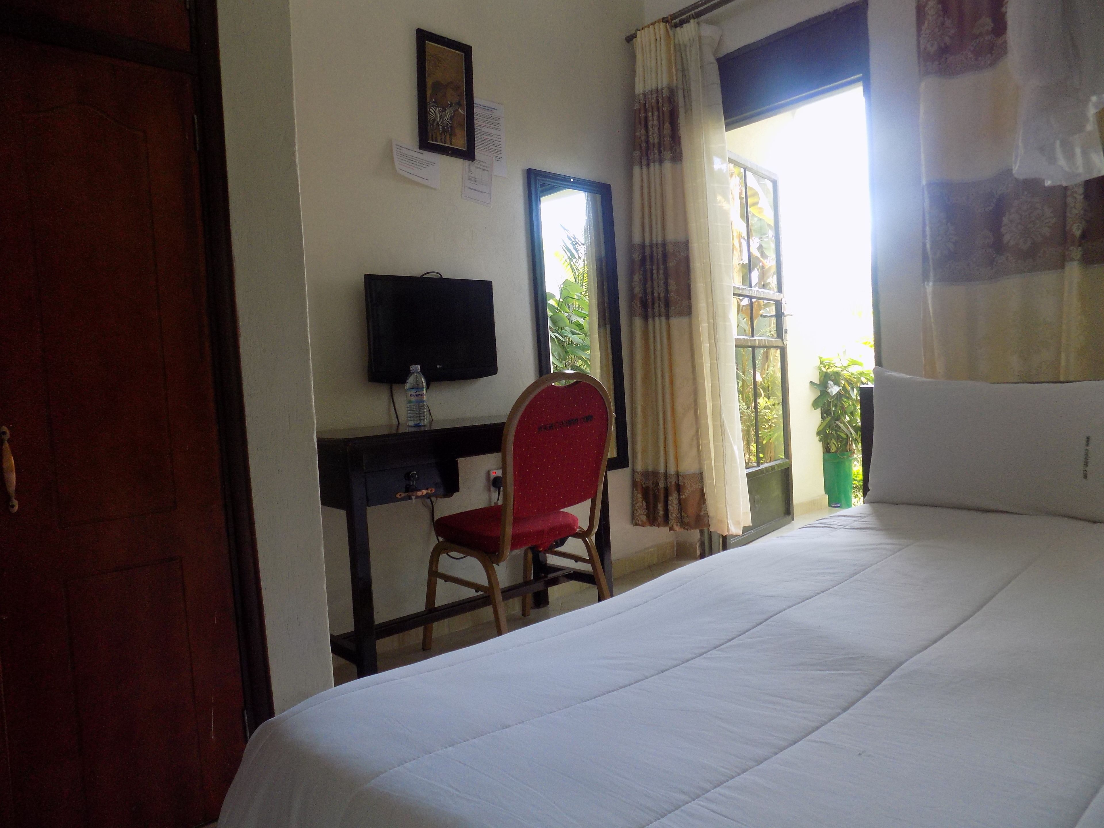 Quarto de hotel com cama, escrivaninha com cadeira, televisão e janela com cortinas.