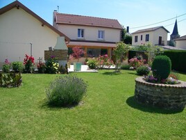Uma casa de dois andares com telhado de telhas, um jardim com diversas plantas e flores, e um vaso de pedra.