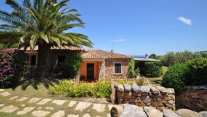 Uma casa de pedra com telhado de cerâmica, rodeada por vegetação exuberante e uma palmeira.