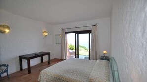 Quarto com cama, escrivaninha e cadeira. Possui janela com cortinas e vista para o verde lá fora.