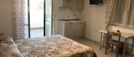 Quarto com cama, escrivaninha, cadeira, pia e televisão na parede.