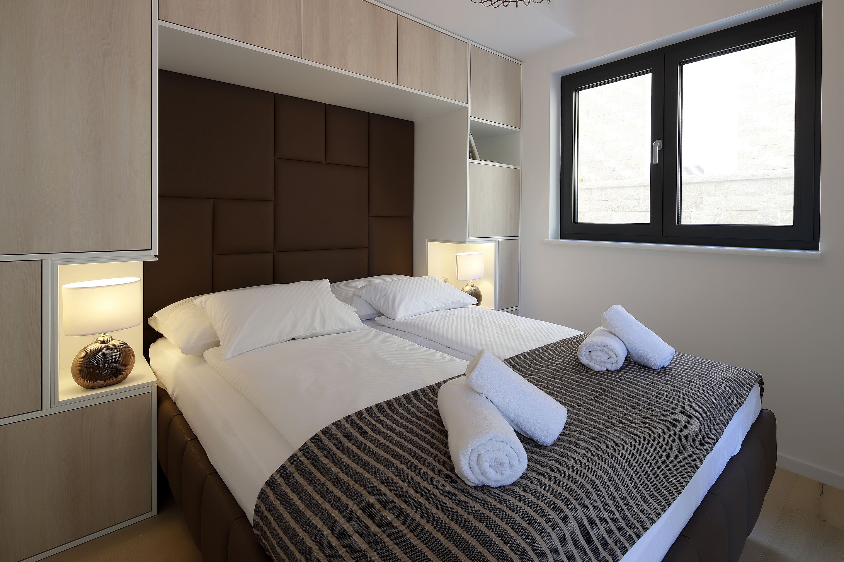 Quarto de hotel com uma cama grande, abajures, janela e espaço de armazenamento embutido.