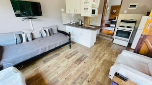 Espaço compacto com sofá, pequena cozinha, televisão na parede e mesa de jantar de madeira.