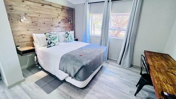 Quarto moderno com cabeceira de madeira, cama grande com colcha cinza e branca, mesa de cabeceira e um banco de madeira.
