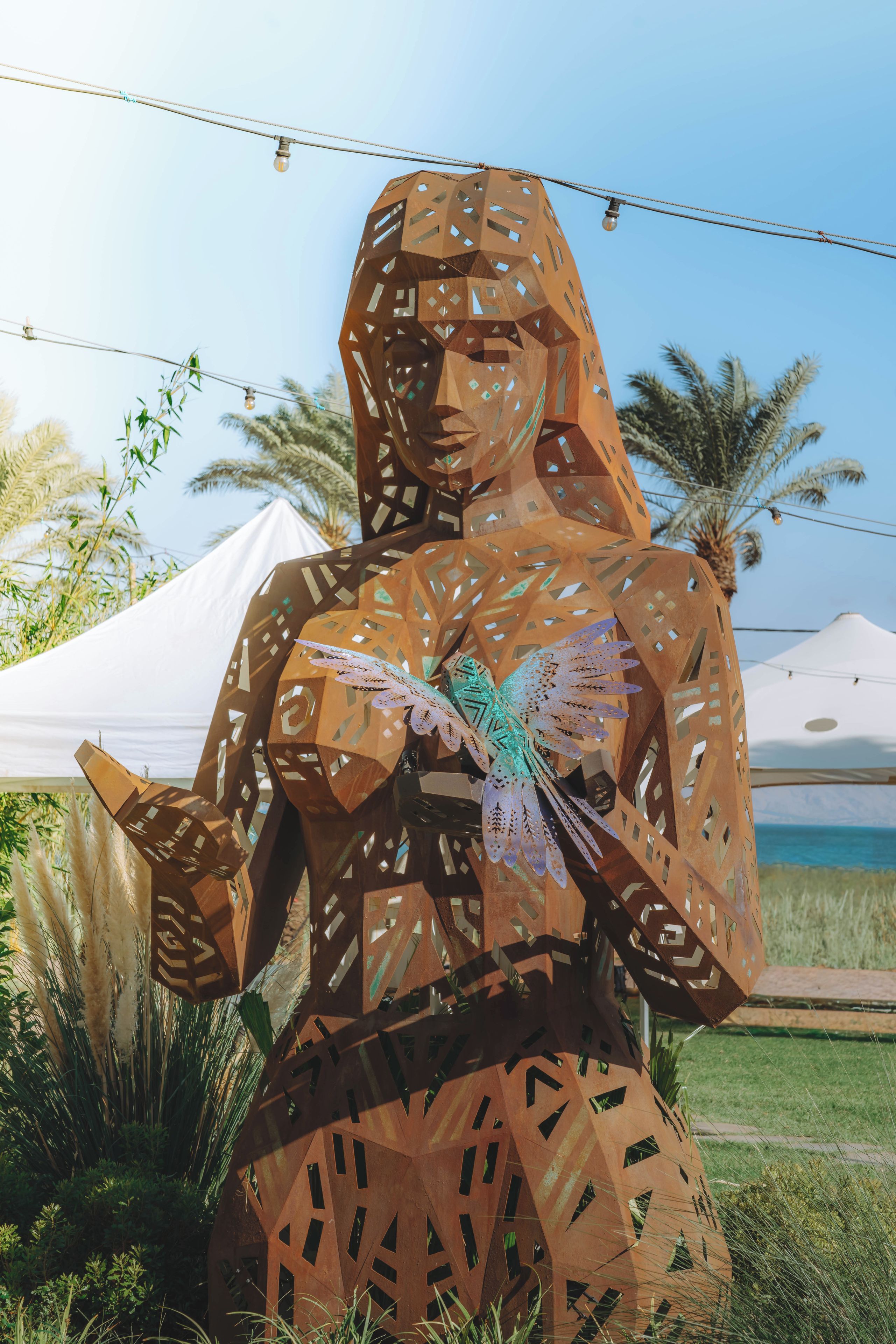 Escultura de uma mulher vestida com manto e cortes geométricos, com palmeiras e céu azul ao fundo.