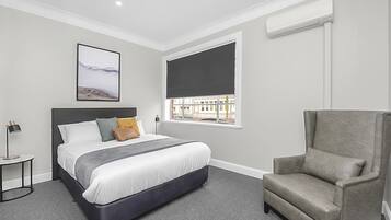 Quarto de hotel moderno com uma cama grande, uma poltrona cinza, uma janela com vista para edifícios e um ar condicionado fixado na parede.