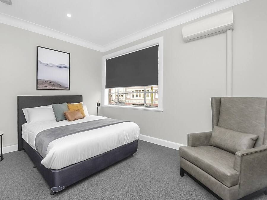 Quarto de hotel moderno com uma cama grande, uma poltrona cinza, uma janela com vista para edifícios e um ar condicionado fixado na parede.