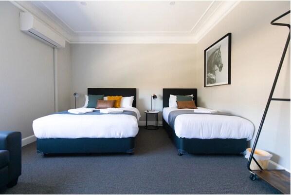 Quarto de hotel com duas camas, uma pequena mesa, uma cadeira, ar-condicionado, um quadro na parede e um abajur.