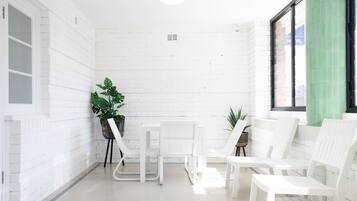 Um quarto claro, com paredes brancas, uma mesa e cadeiras, um vaso com planta e uma janela com vista para um edifício.