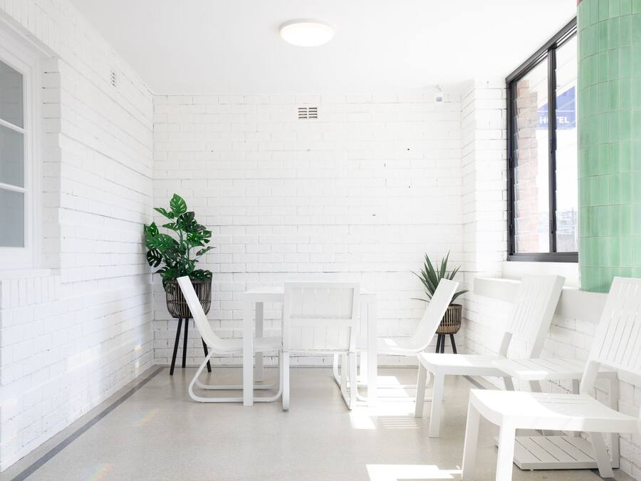 Um quarto claro, com paredes brancas, uma mesa e cadeiras, um vaso com planta e uma janela com vista para um edifício.