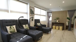 Um quarto com três sofás e pufes escuros dispostos em linha, uma mesinha de apoio com abajur e uma estante decorativa com plantas e um busto.