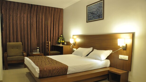 Quarto de hotel com uma cama grande, cabeceira de madeira, duas mesas de cabeceira, uma cadeira e uma cortina.