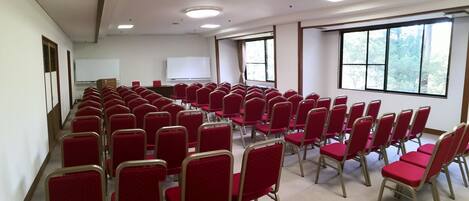 Uma sala de conferências com fileiras de cadeiras vermelhas, um quadro branco e janelas amplas.